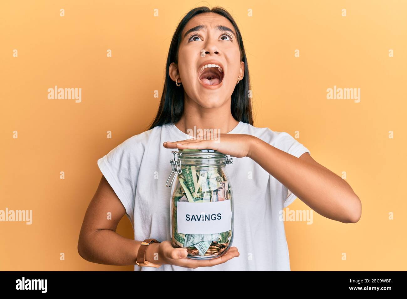Young asian woman holding jar with savings angry and mad screaming ...