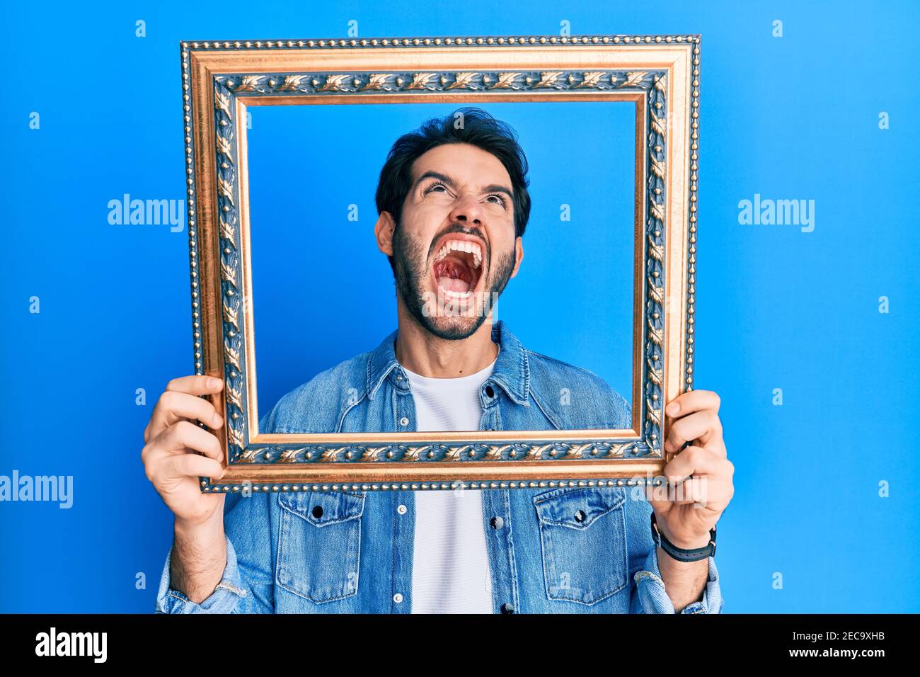 Young hispanic man holding empty frame angry and mad screaming ...