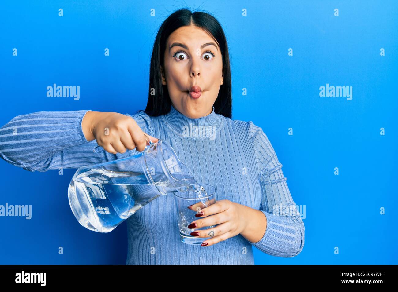 Beautiful brunette woman pouring water in glass making fish face with mouth and squinting eyes ...