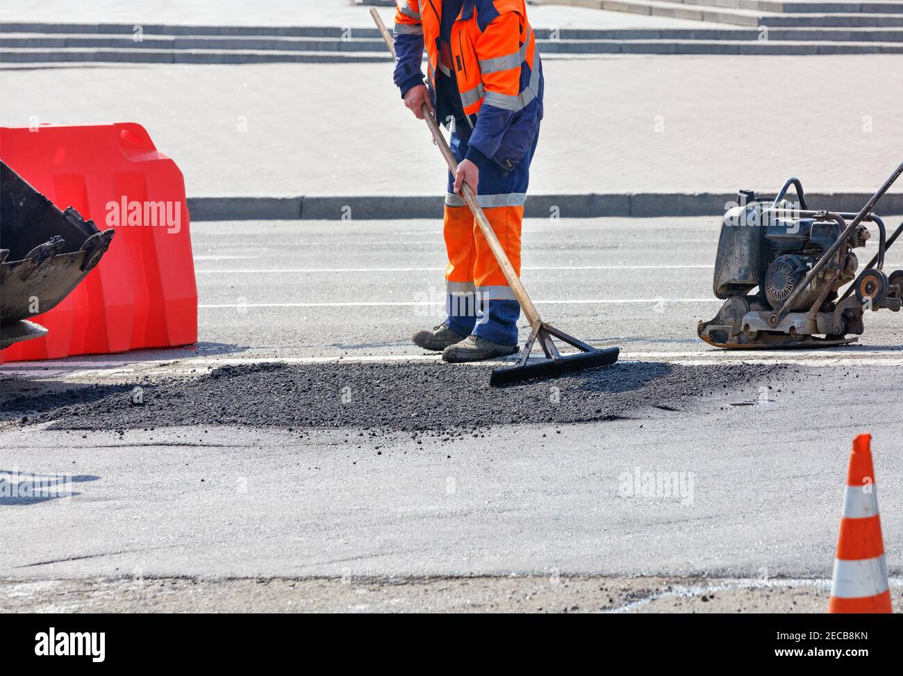 A road worker in a bright orange overalls levels fresh asphalt with a ...