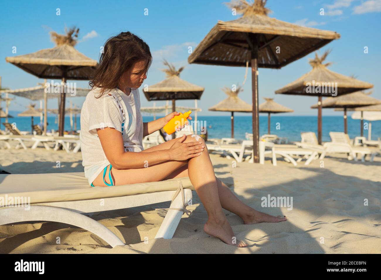 Mature Woman Sitting Onsun Lounger On Sandy Beach Applying Sunscreen To