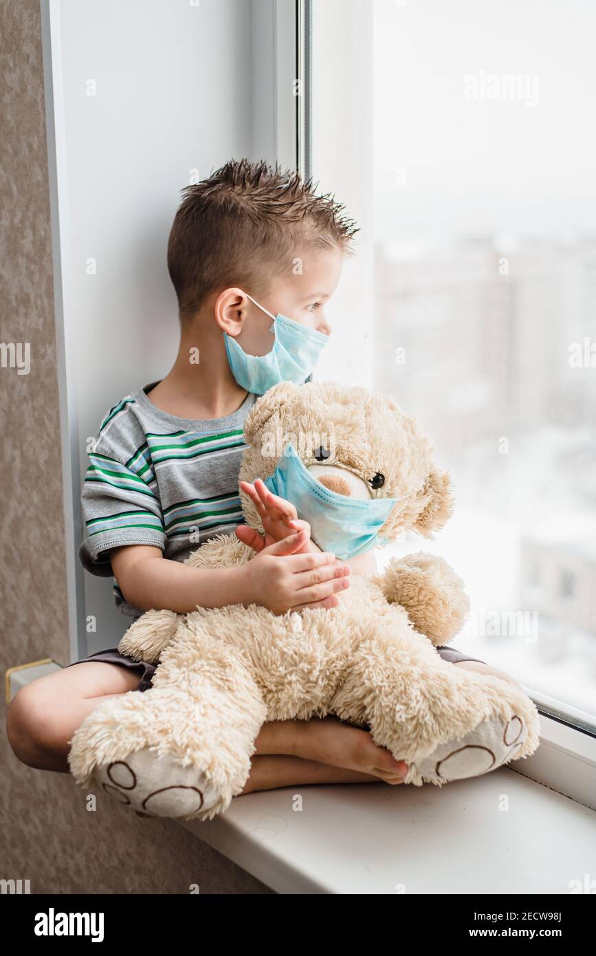 a little boy in a mask sits at home in quarantine and looks out the ...