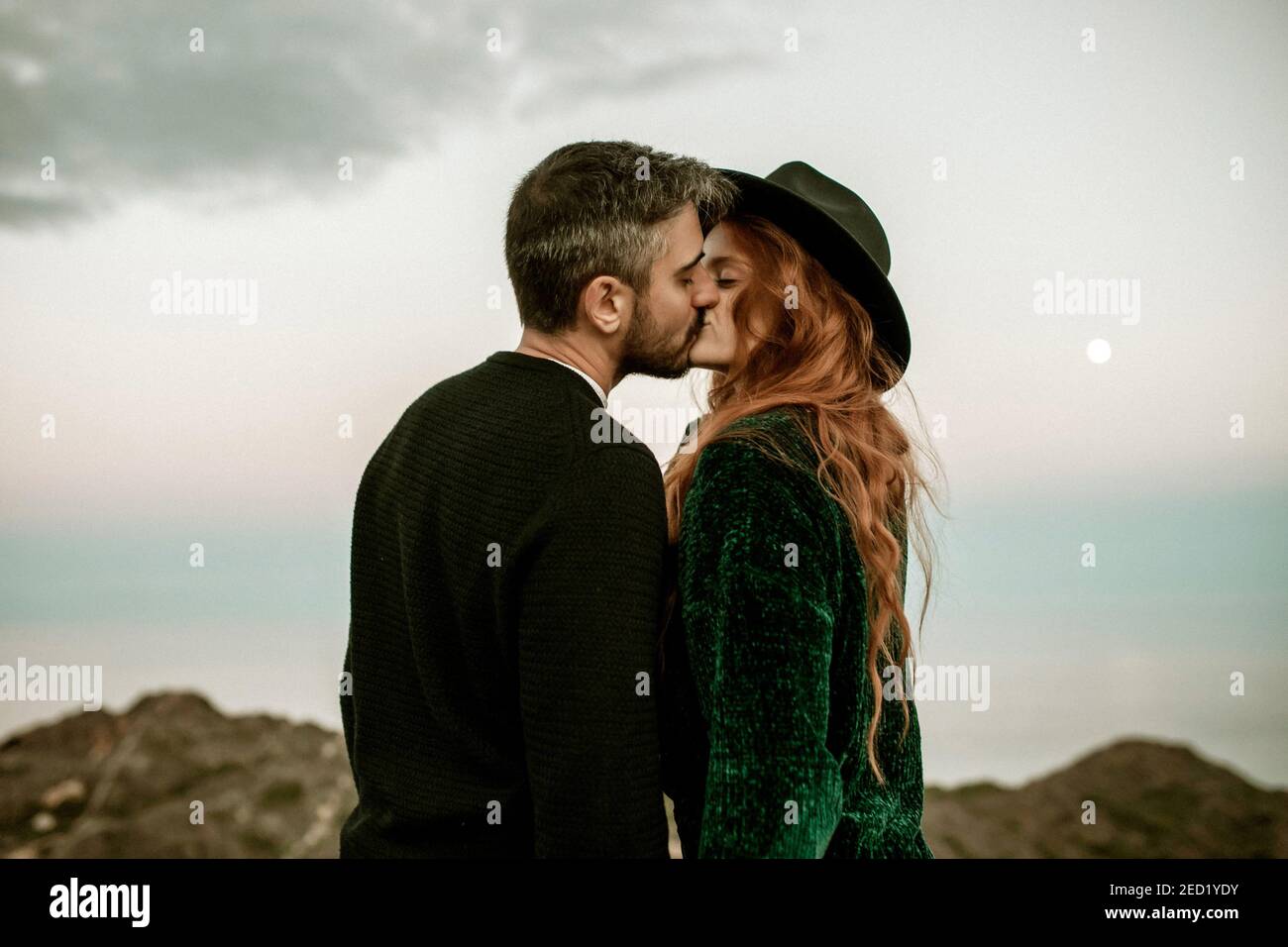 Side view of calm couple in stylish clothes and hat standing kissing at ...