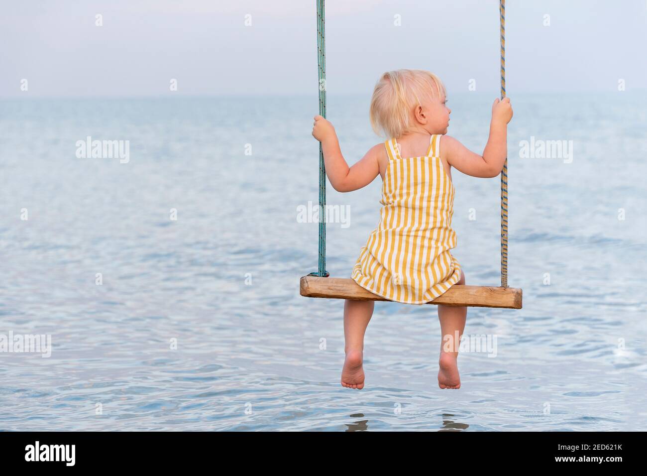 Blonde girl in yellow dress sitting on a rope swing over water. Swing ...