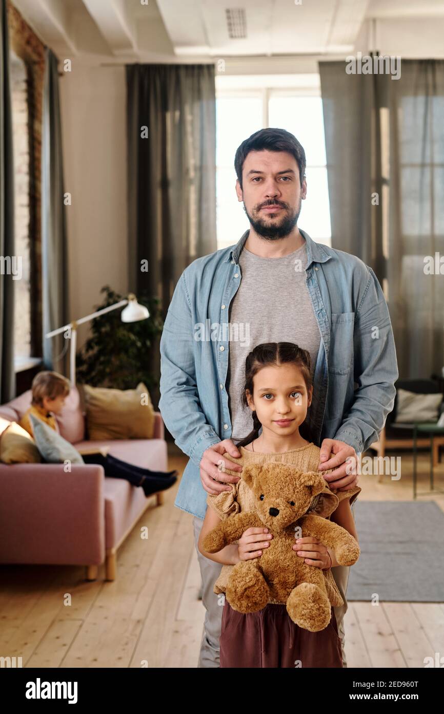 Serious young man standing by his little pretty daughter with soft ...