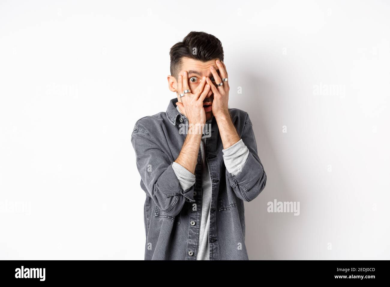 Scared young man peeking through fingers with nervous face, staring at ...