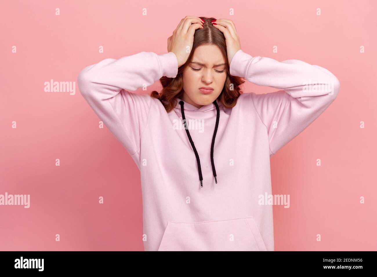 Portrait of sick teenage girl in hoodie clasping hands over sore head ...