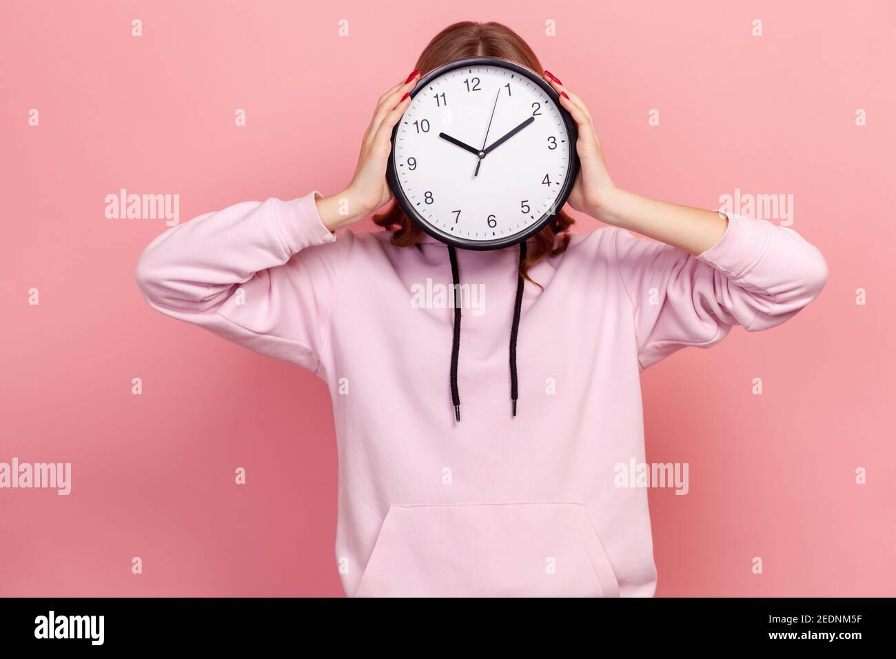 Portrait of young brunette female in hoodie holding wall clock hiding ...