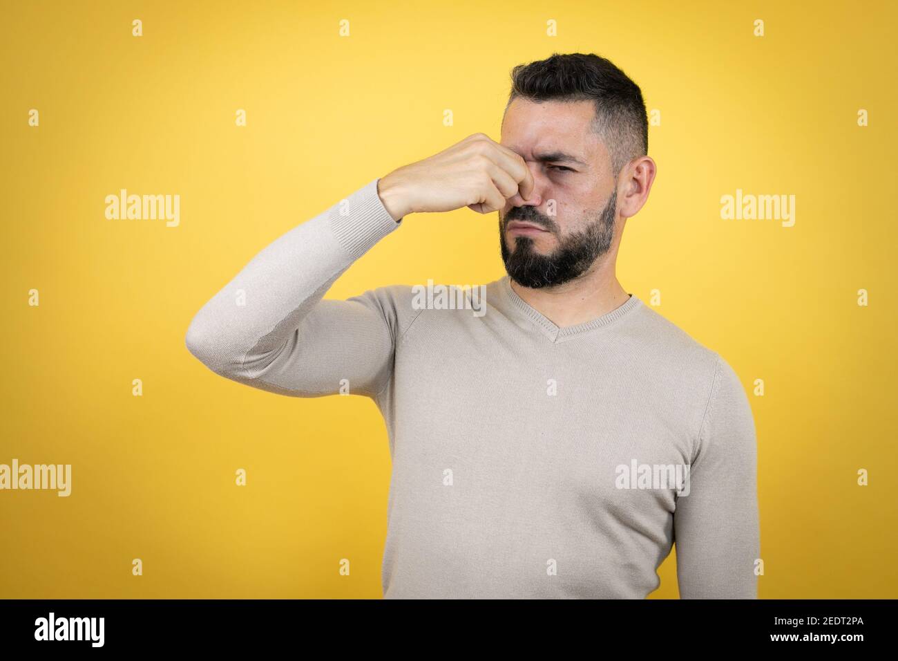 Handsome man with beard wearing sweater over yellow background smelling ...