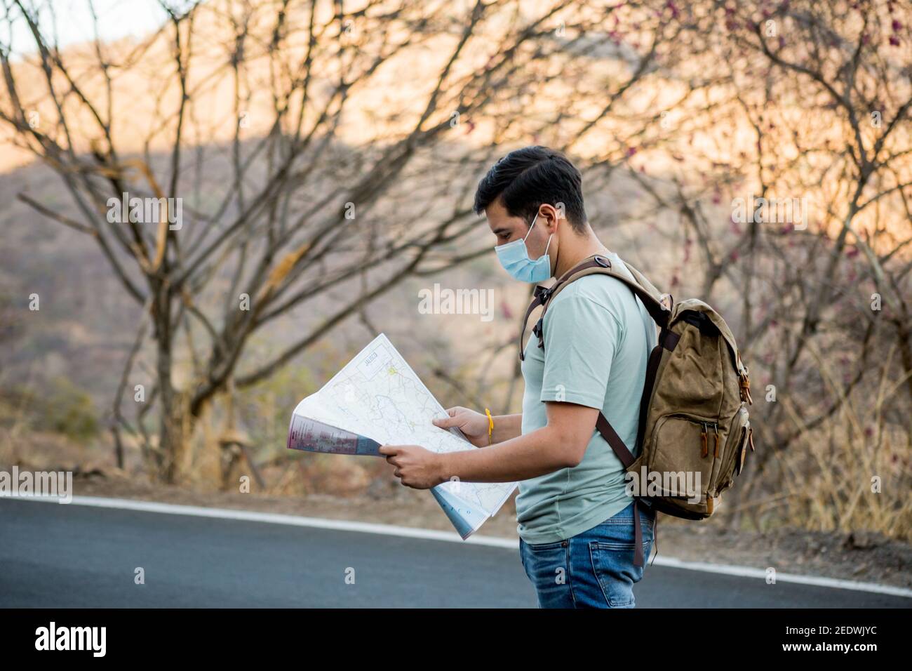 Male traveler with a backpack and maps wearing a sanitary mask standing ...