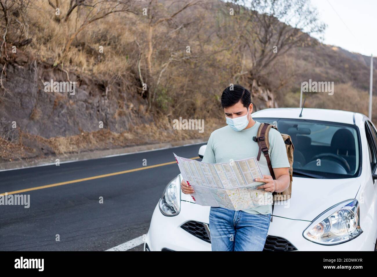 Male traveler wearing a sanitary mask leaning on his car looking at ...
