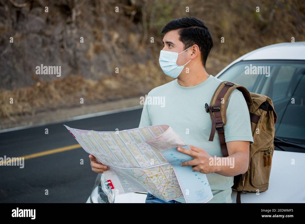Male traveler wearing a sanitary mask leaning on his car holding maps ...