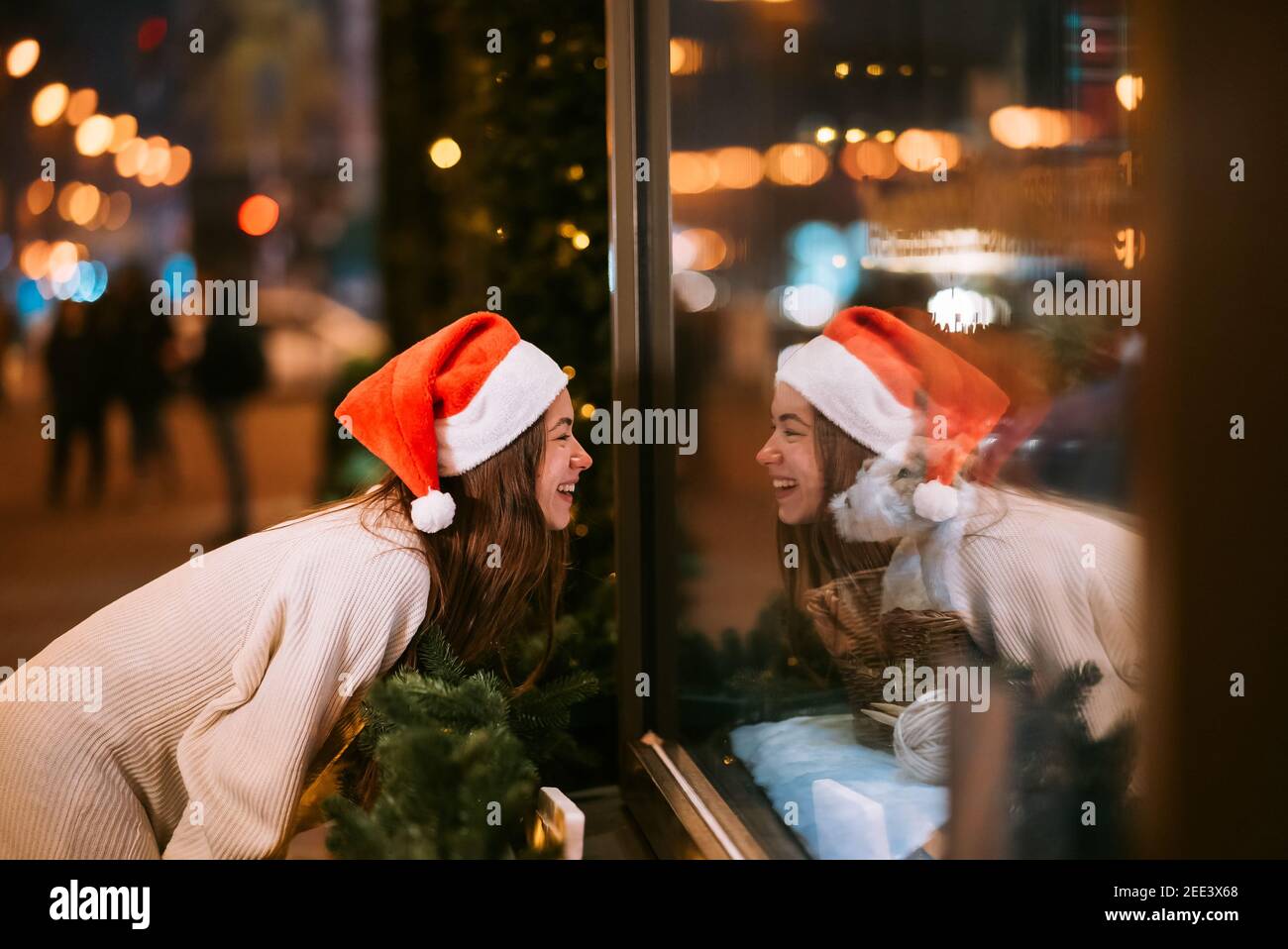 Smiling girl looking through shop window, city lights on background ...