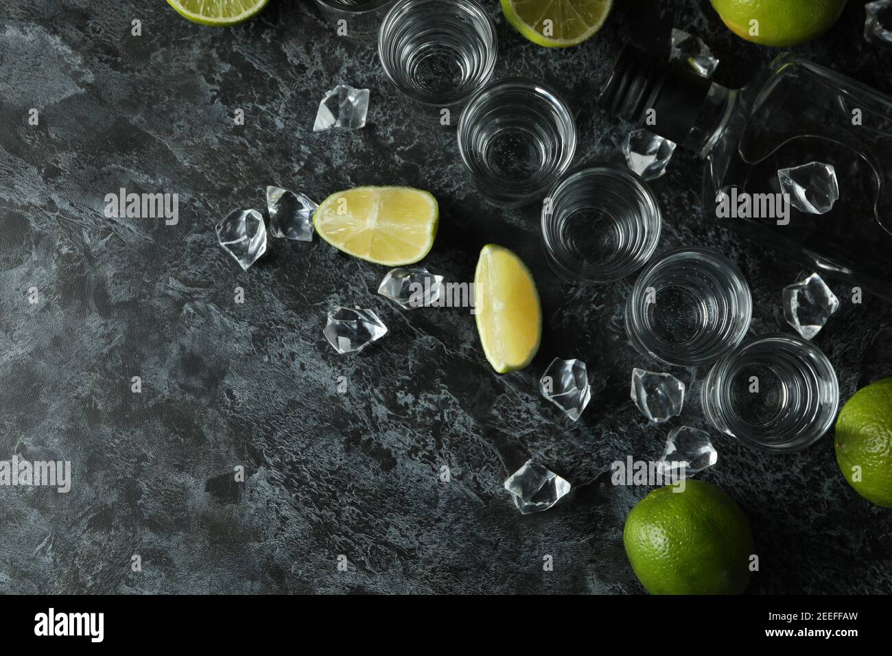 Bottle of vodka, shots, ice cubes and lime slices on black smokey table ...