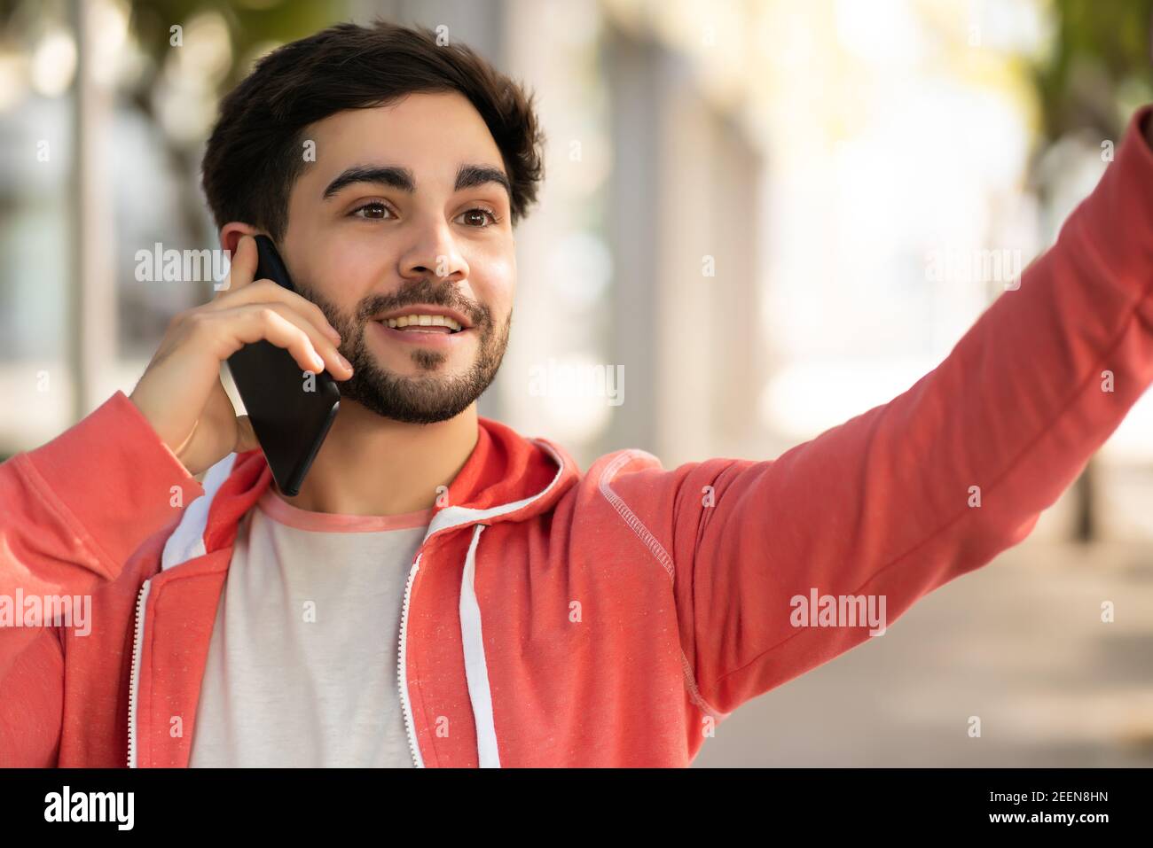 Portrait of young man talking on the phone and raising his hand to hail ...