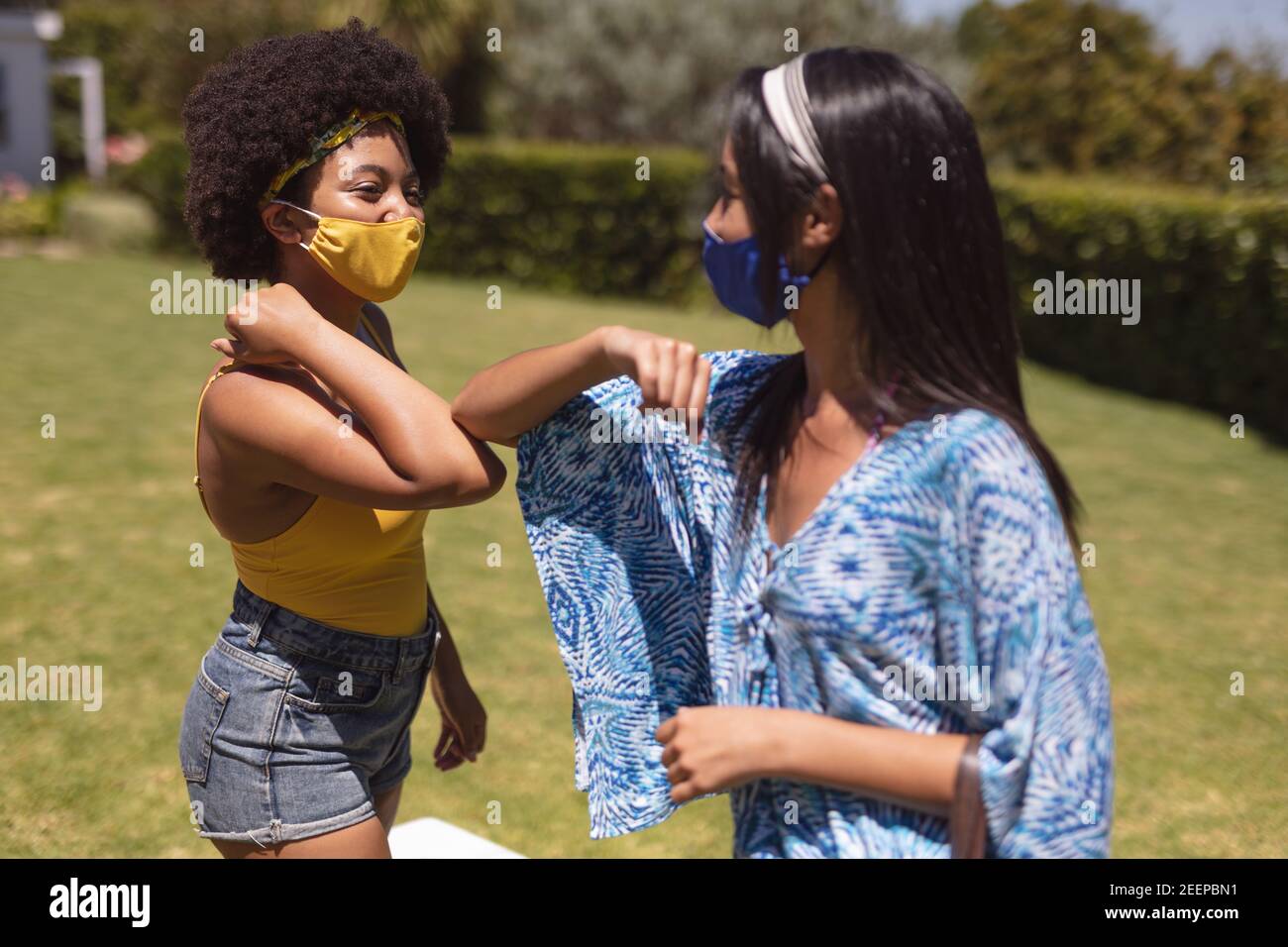Two diverse female friends wearing face masks bumping elbows at a pool ...