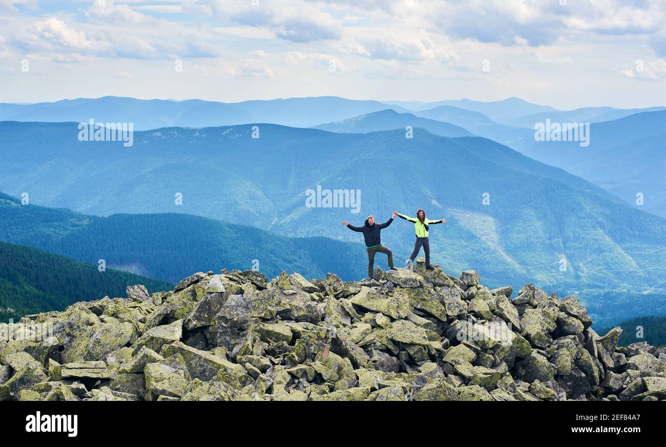 Happy couple holding hands up, standing on top of cliff, beautiful mountains scenery on ...