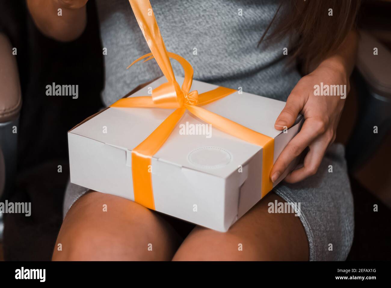Selective focus shot of a lady in a short dress opening a white gift box with an oran ribbon ...
