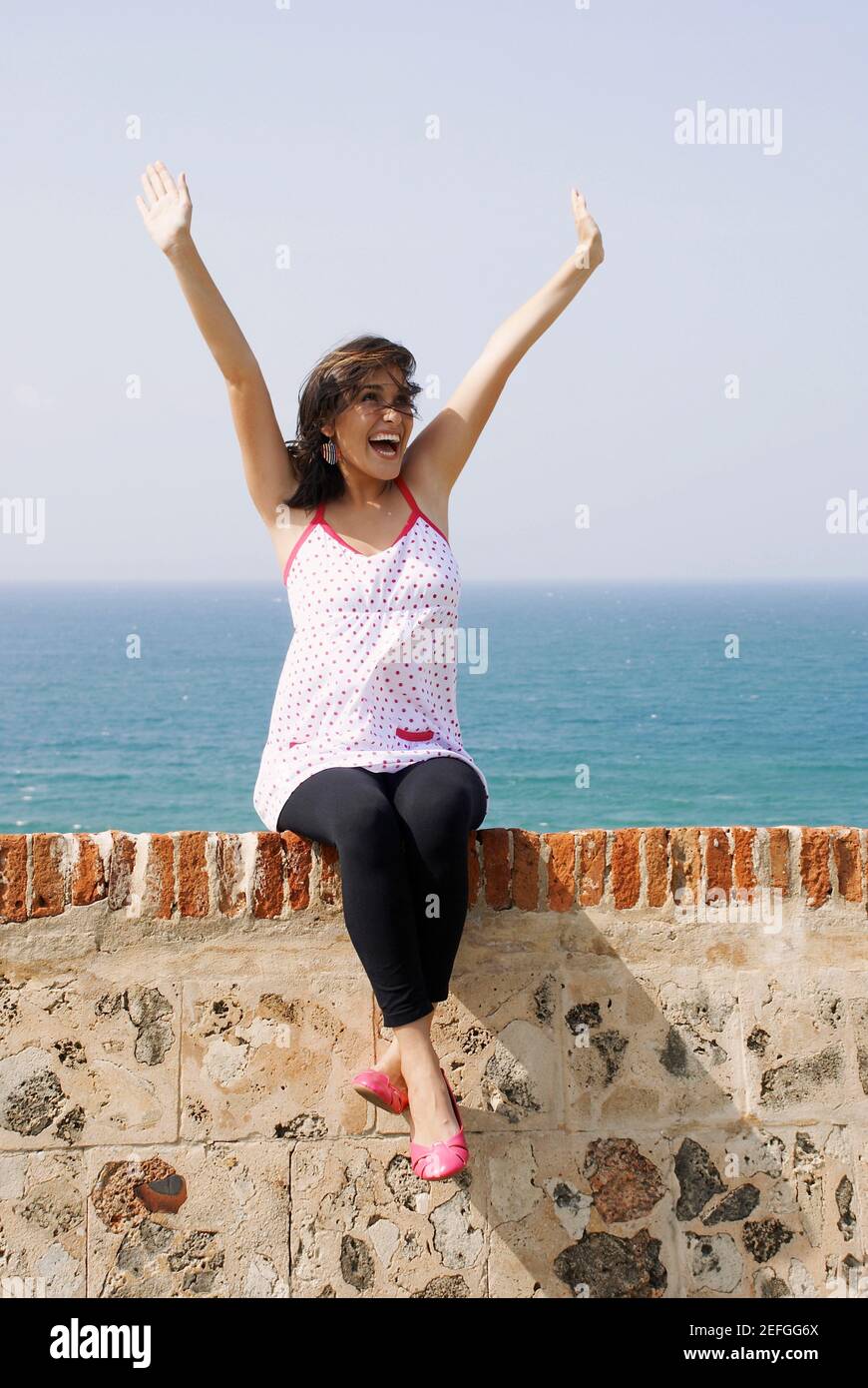 Young woman sitting on the ledge with her arms raised and laughing ...