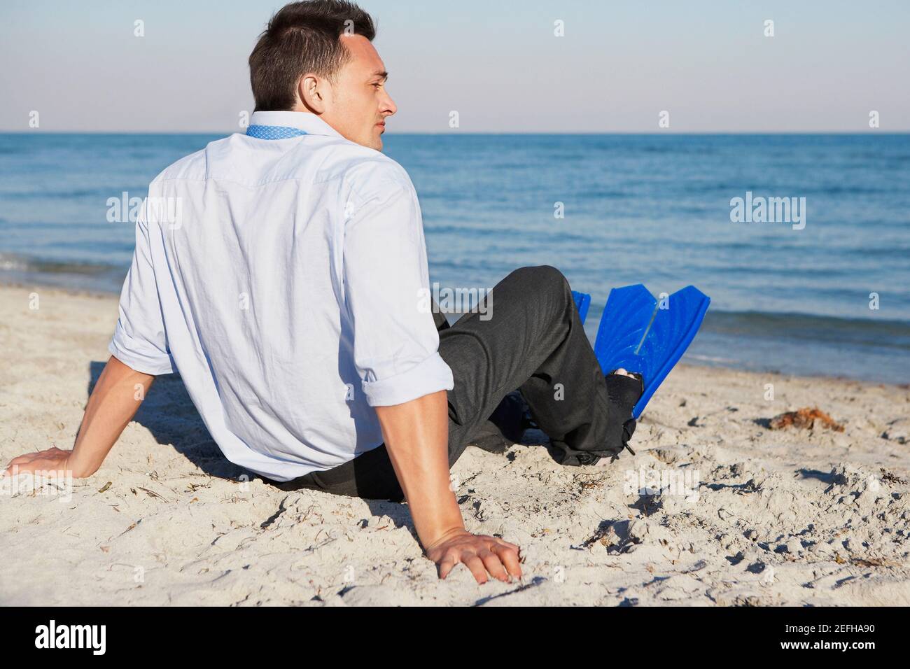 Rear view of a young man wearing flippers and sitting on the beach ...