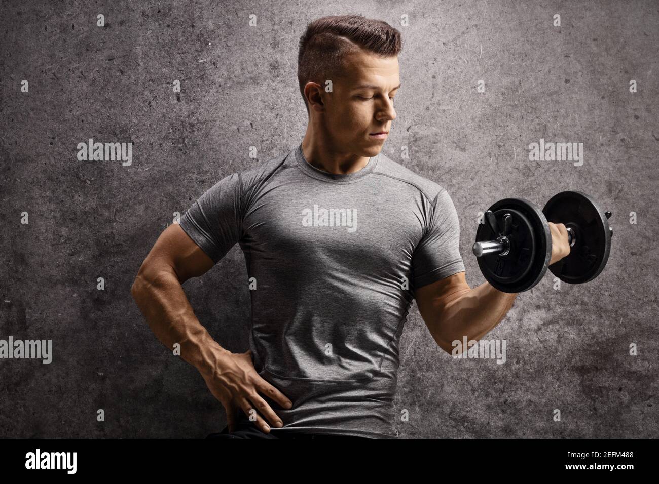Bodybuilder exercising with a dumbbell and leaning on a rusty gray wall ...