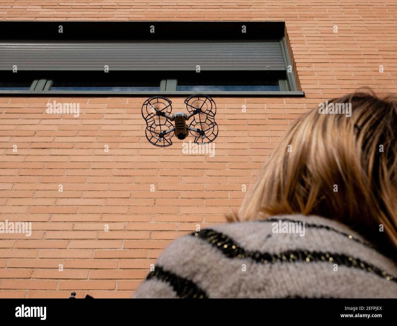 A woman piloting a drone with propeller guard flying against a brick ...