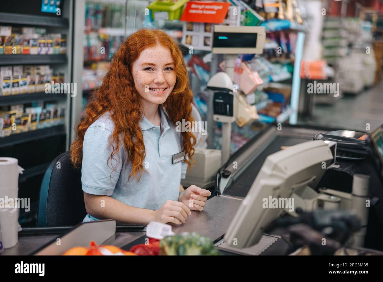 Portrait of a young woman working at grocery store checkout counter. Female cashier sitting ...