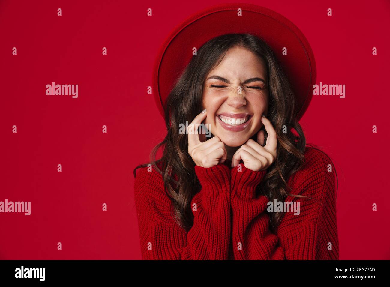 Joyful beautiful girl in hat pointing fingers at her cheeks and ...