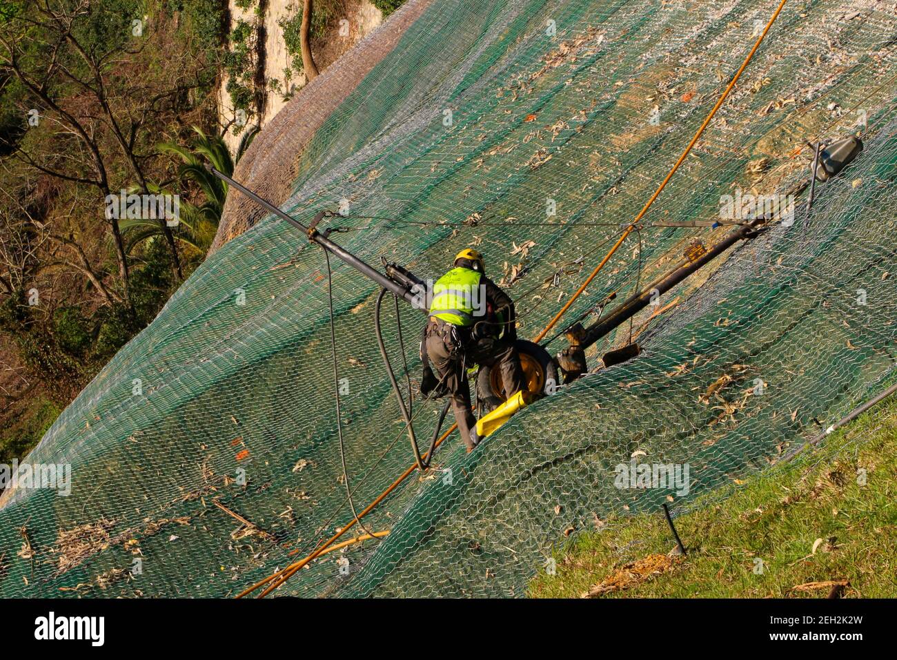 Two men tethered by ropes using a drill rig to insert soil nails to ...
