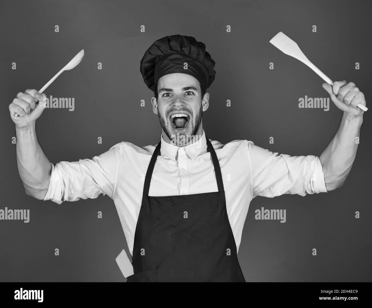 Chef with excited face holds wooden spoon and spatula on red background ...