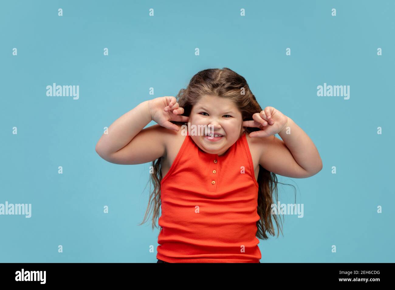 Closing ears. Happy, smiley little caucasian girl isolated on blue ...