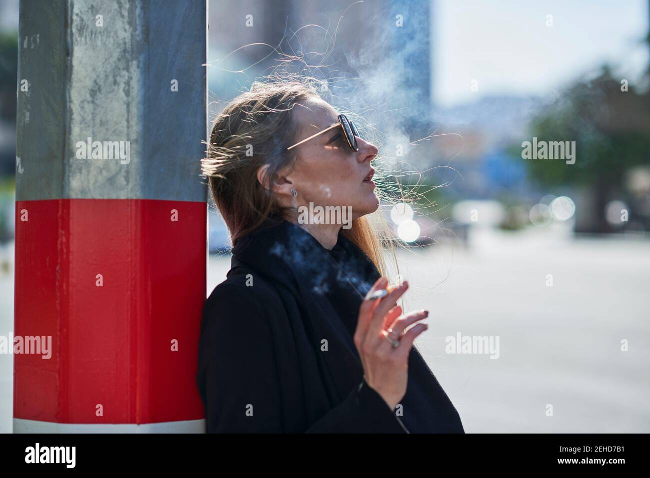 Side view of young female in scarf smoking cigarette near post on urban ...