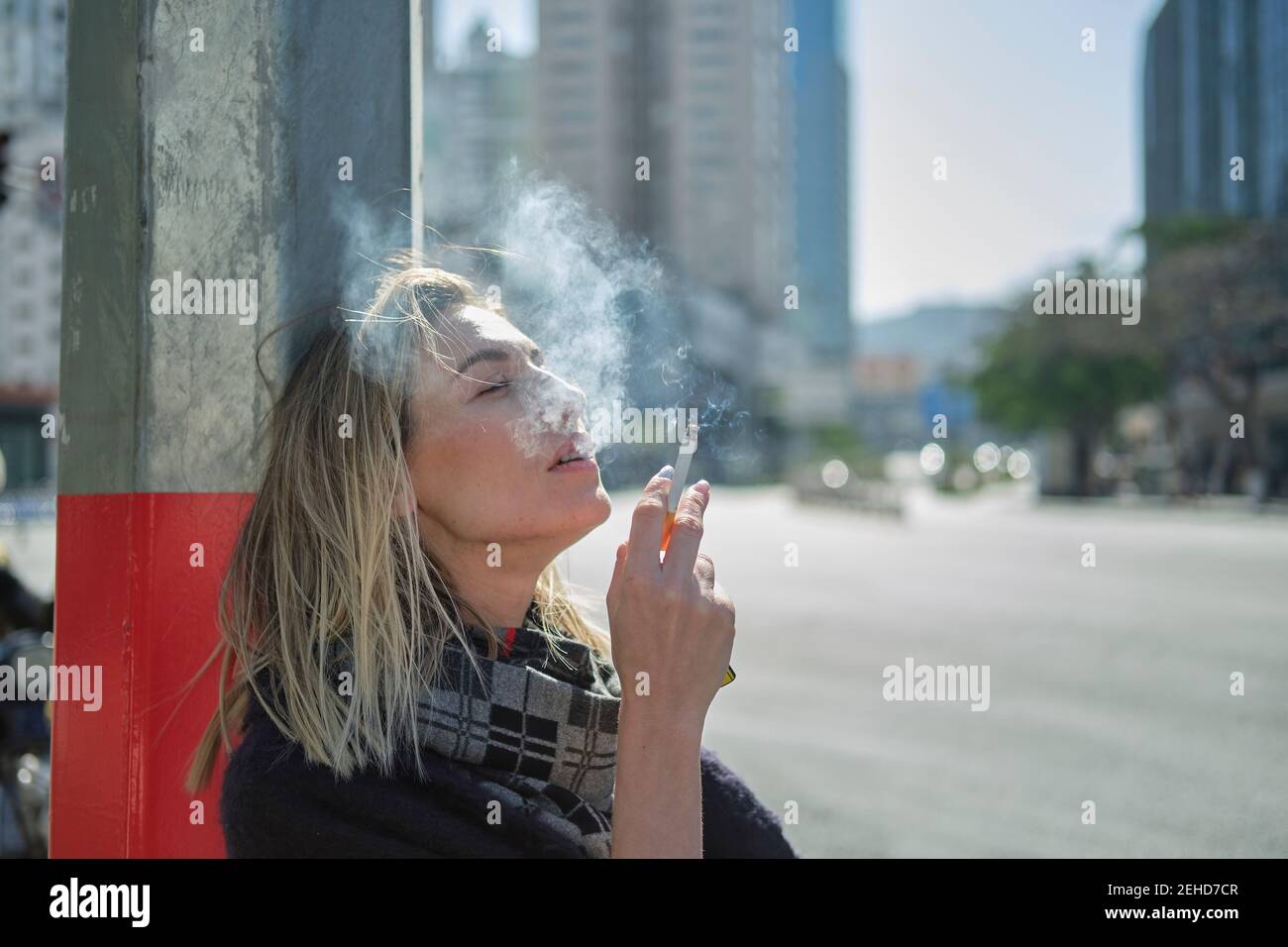Side view of young female in scarf smoking cigarette near post on urban ...