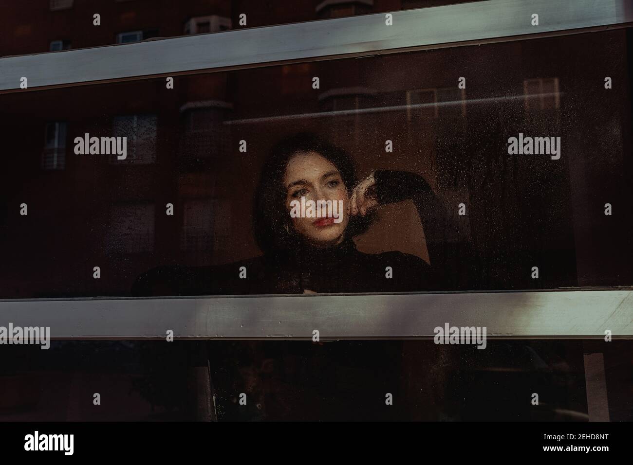 Through glass of thoughtful female standing near window and observing ...