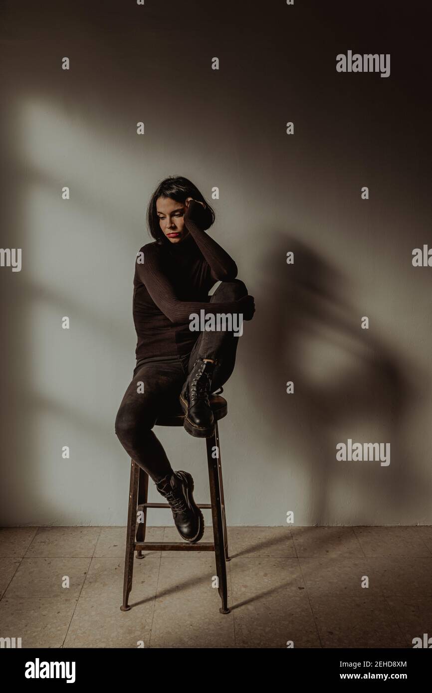 Slim female in black outfit sitting on wooden stool with raised legs in ...