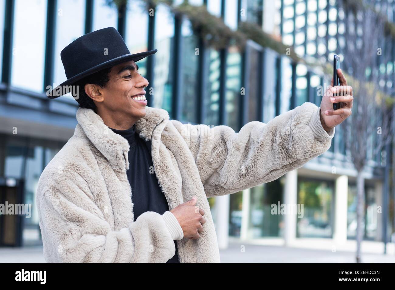 Cheerful ethnic male wearing trendy outerwear and hat smiling widely ...