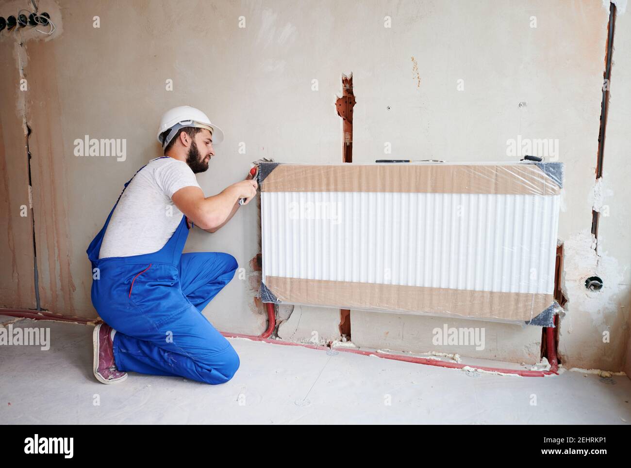 Young plumber, wearing blue overalls, white helmet, installing water ...