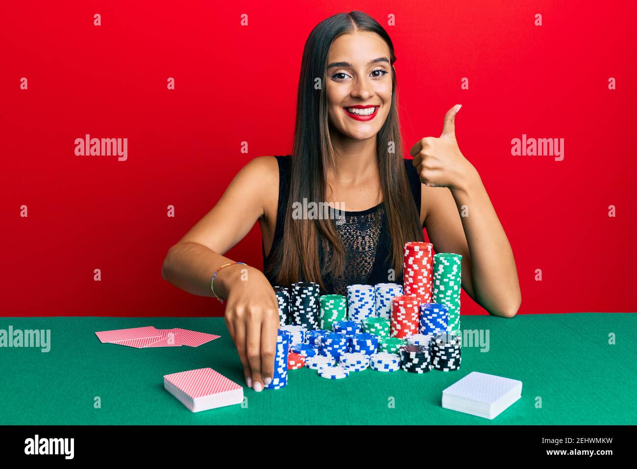 Young hispanic woman sitting on the table holding casino chips smiling happy and positive, thumb ...