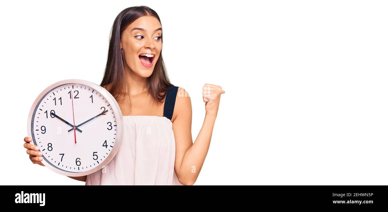 Young hispanic woman holding big clock pointing thumb up to the side ...