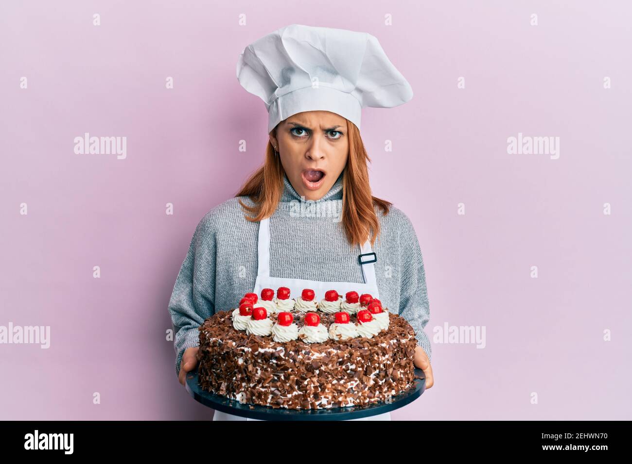 Hispanic young chef woman holding chocolate cake in shock face, looking ...