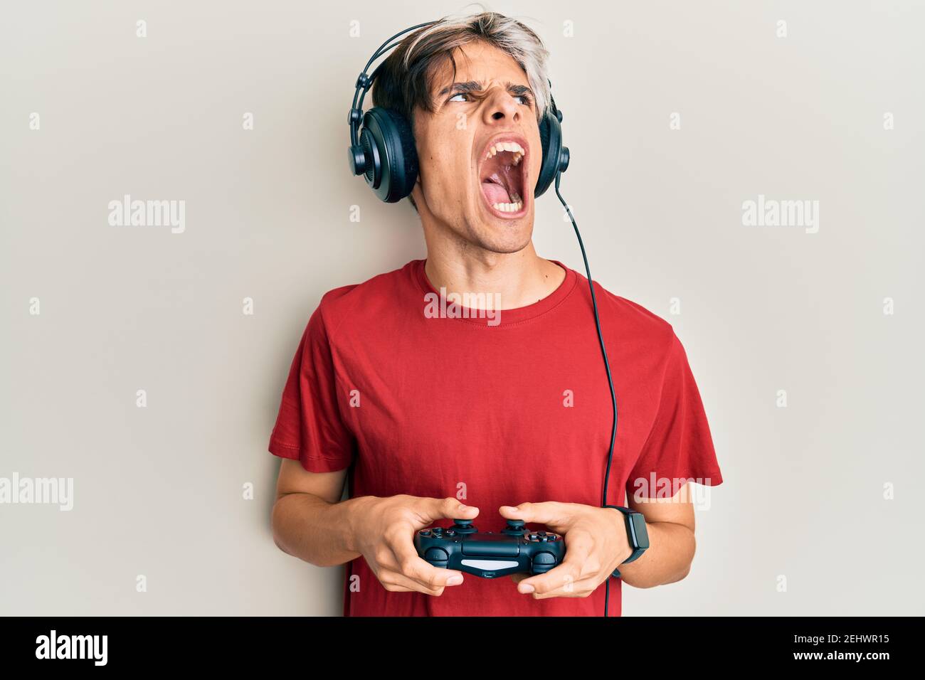Young hispanic man playing video game holding controller angry and mad ...