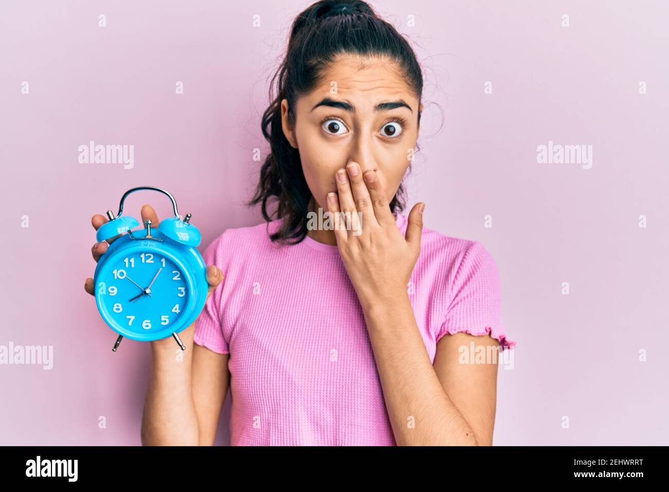 Hispanic teenager with dental braces holding alarm clock covering mouth ...