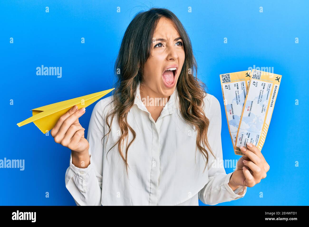 Young brunette woman holding paper airplane and boarding pass angry and ...