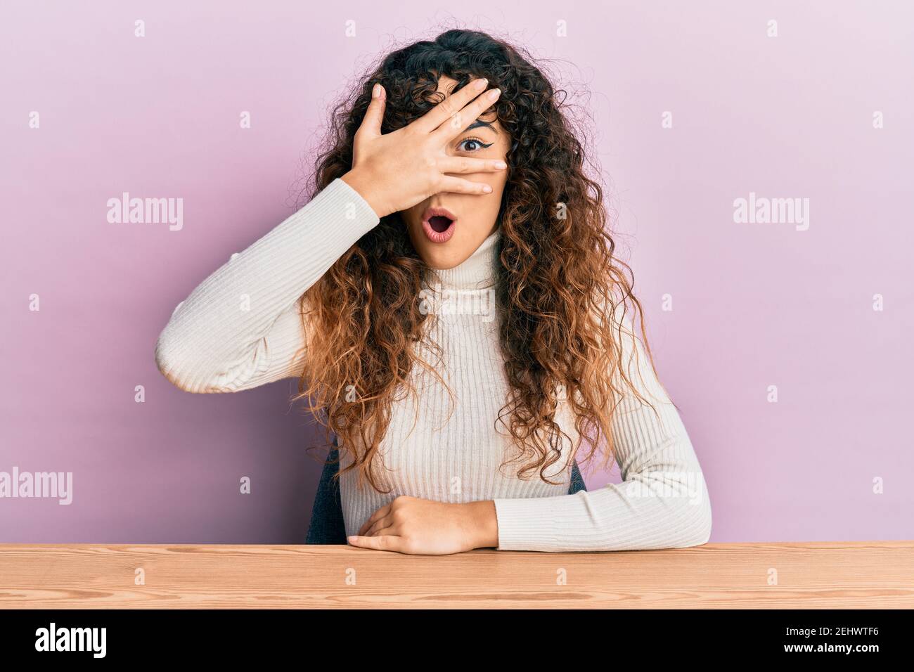 Young hispanic girl wearing casual clothes sitting on the table peeking ...