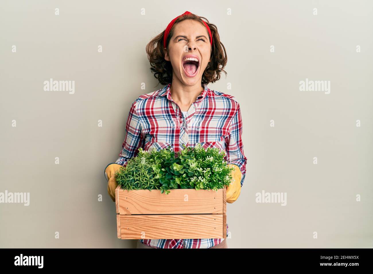 Young brunette woman wearing gardener clothes holding wooden plant pot ...