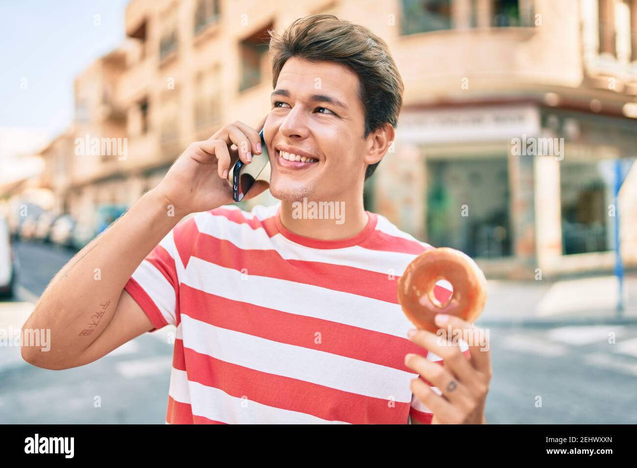 Young caucasian man talking on the smartphone eating donut at the city ...