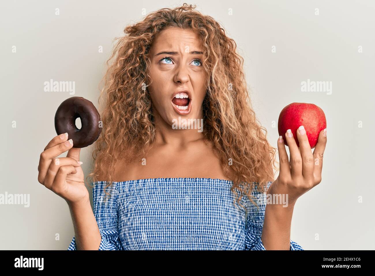 Beautiful caucasian teenager girl holding red apple and donut angry and ...