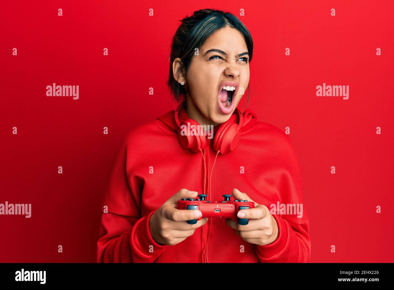 Young latin woman playing video game holding controller angry and mad ...