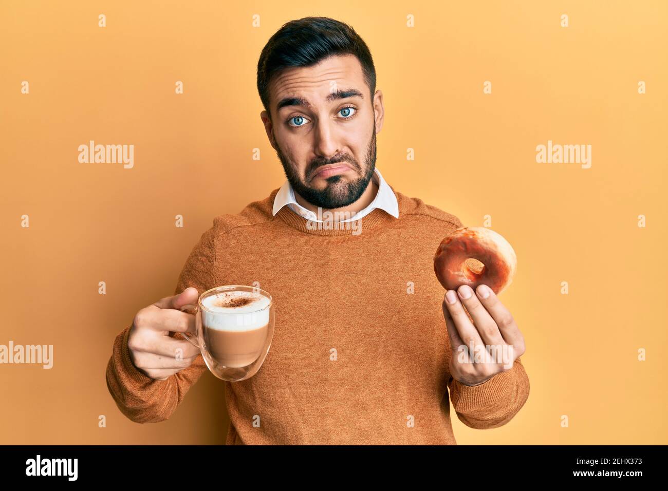 Young hispanic man eating doughnut and drinking coffee depressed and ...