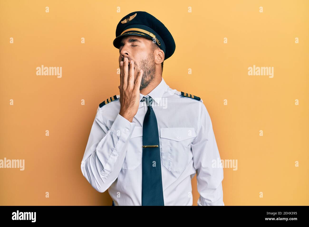 Handsome hispanic man wearing airplane pilot uniform bored yawning ...